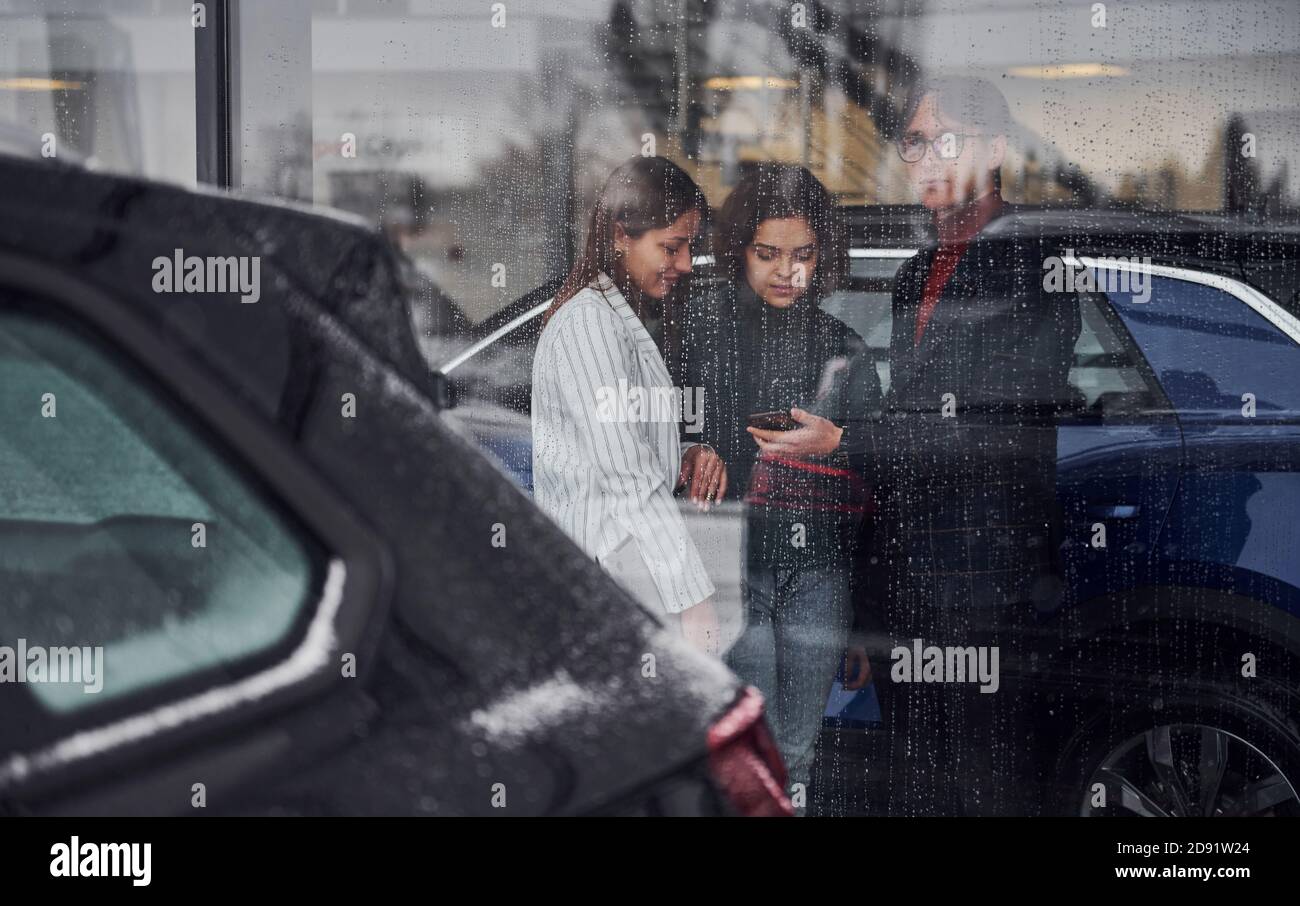 Friends standing behind glass at rainy day. Girl holds laptop in hands ...