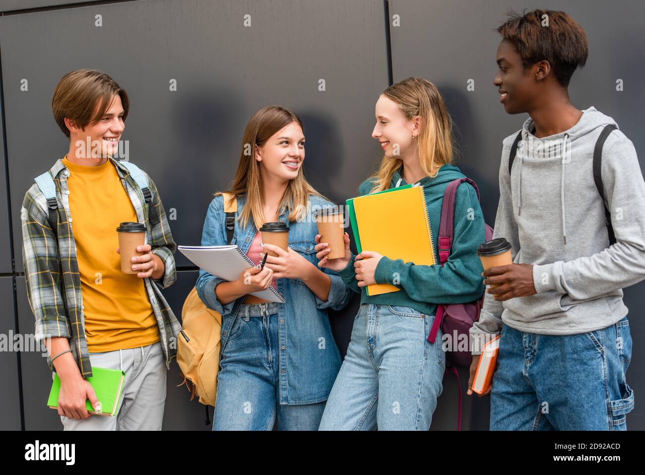 Positive teenagers with notebooks and coffee to go standing near ...