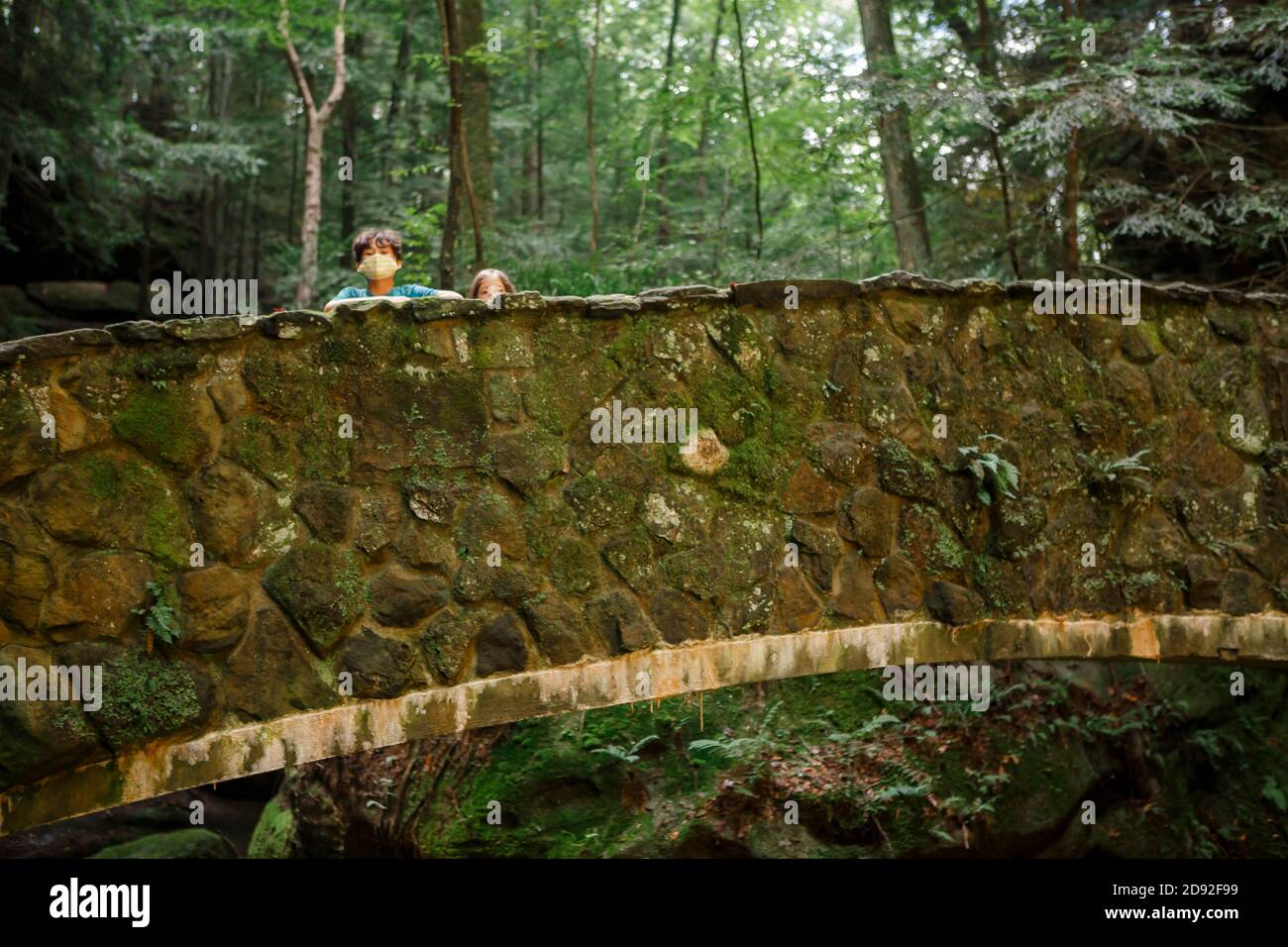 Two small children peer their heads over stone bridge in the forest ...