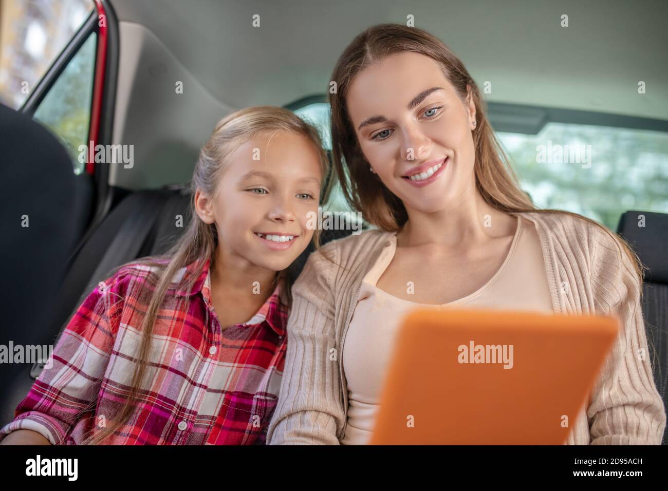 Smiling daughter and her mom checking something on tablet on backseat ...