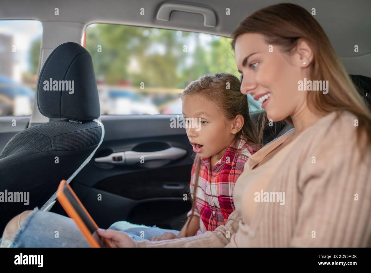 Surprised daughter and her mom checking something on tablet on backseat ...