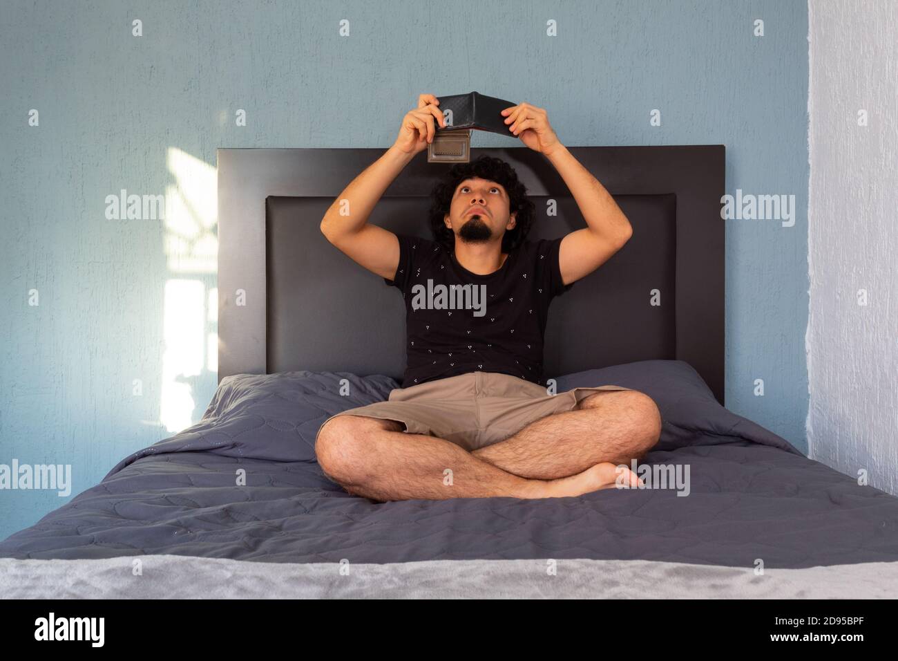 Hispanic young man sitting on a bed looking at his wallet with no money ...