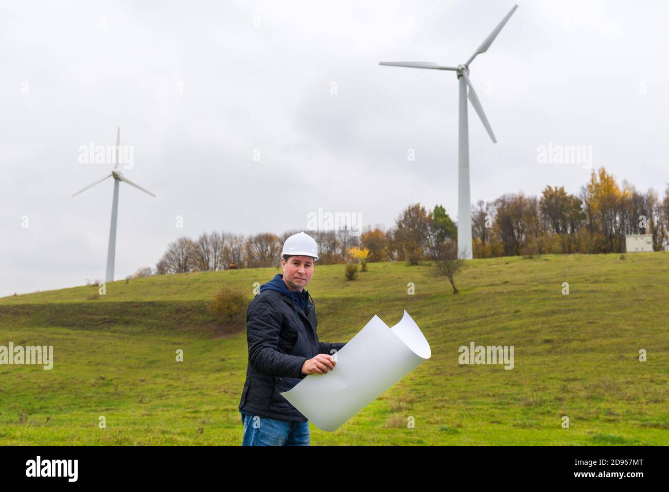 Side view portrait of one male engineer holding a plan at wind turbine at autumn cloudy sky ...