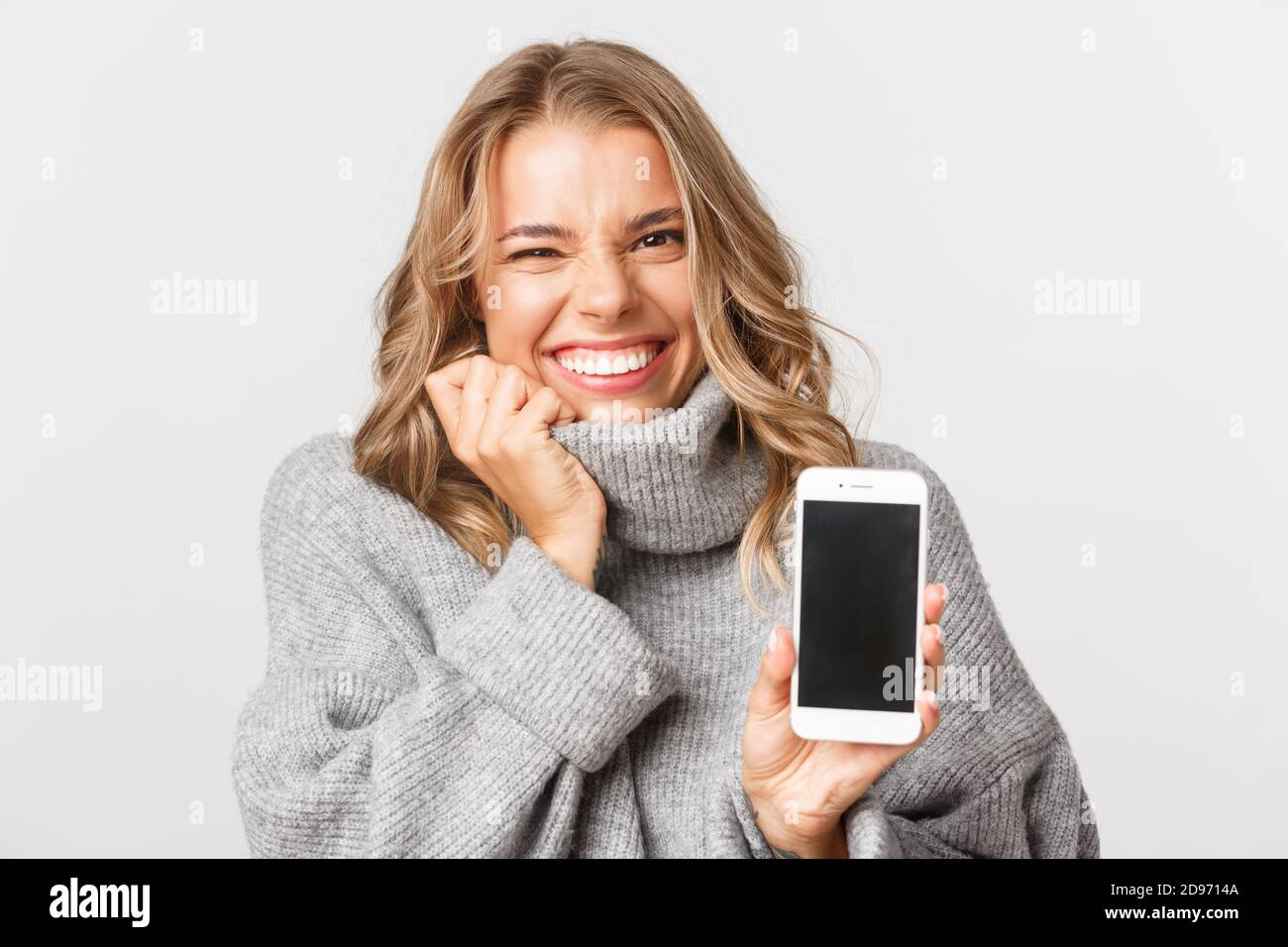 Close-up of happy and excited blond girl, smiling while showing mobile ...