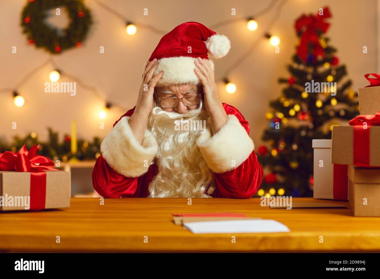 Sad old bearded Santa Claus sitting at a table holding his head ...