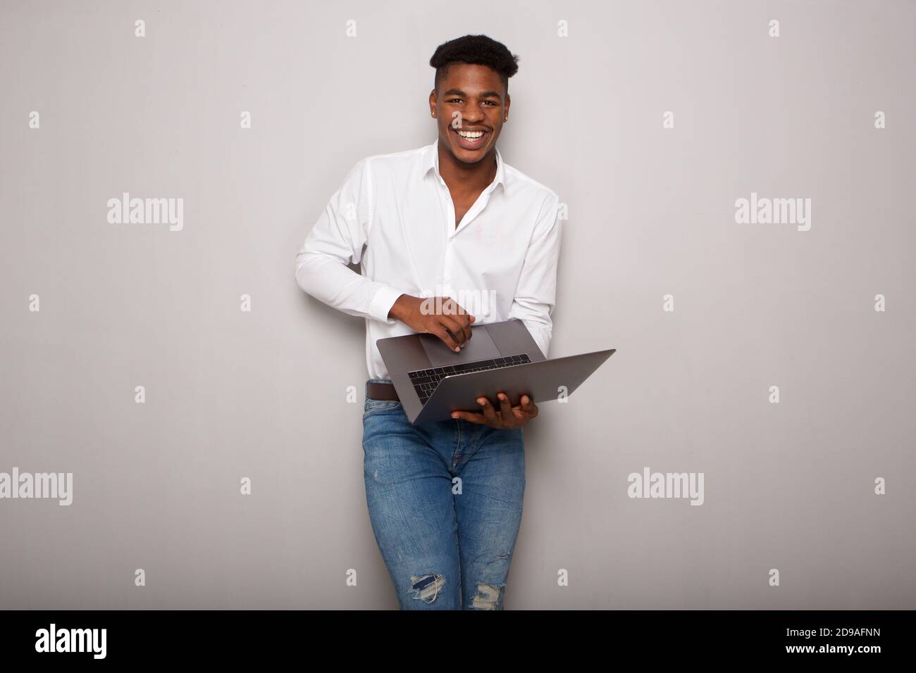 Portrait of happy young black man holding laptop computer by gray wall ...