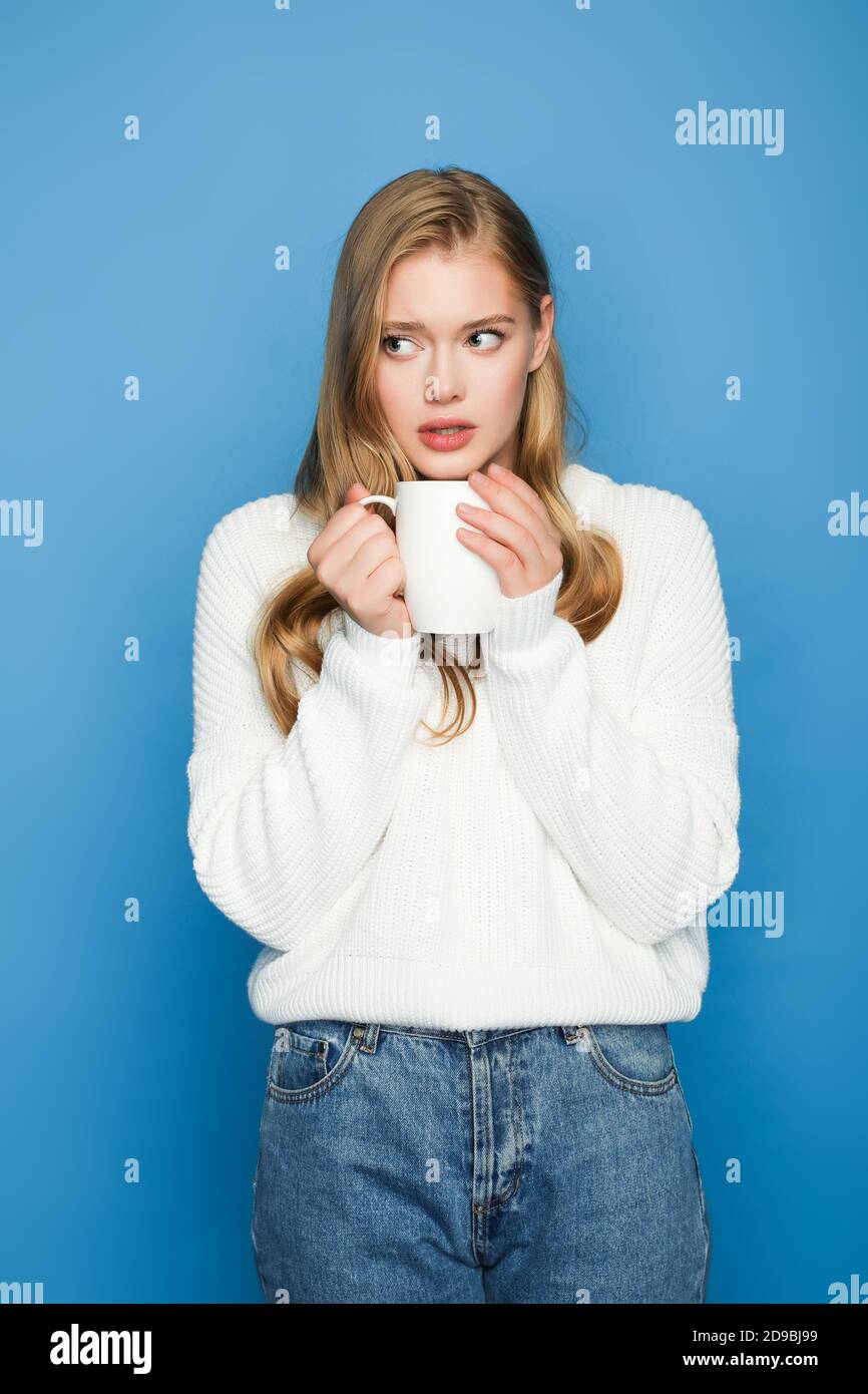 worried blonde beautiful woman in sweater with mug isolated on blue ...
