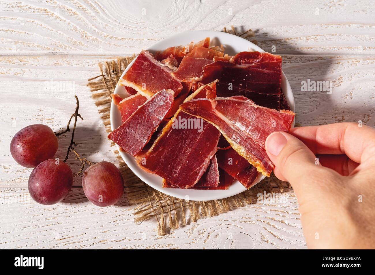 Woman hand takes a slice of jamon from a plate over white wood table ...