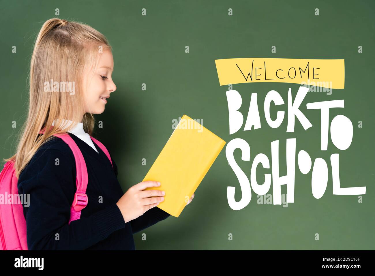 side view of schoolgirl reading book near green chalkboard with welcome ...