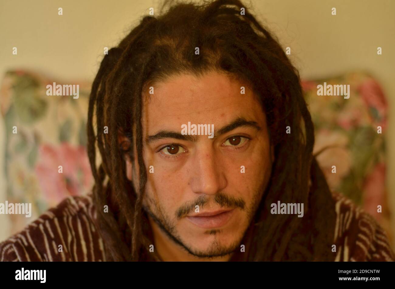Spanish man with deadlocks sitting on a colorful couch and looking into ...