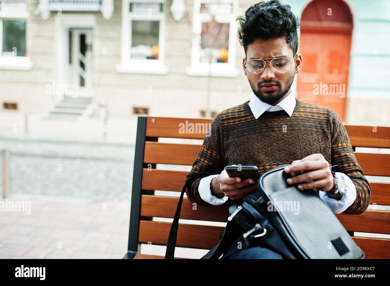 Portrait of young stylish indian man model pose in street, sitting on bench with handbag and ...