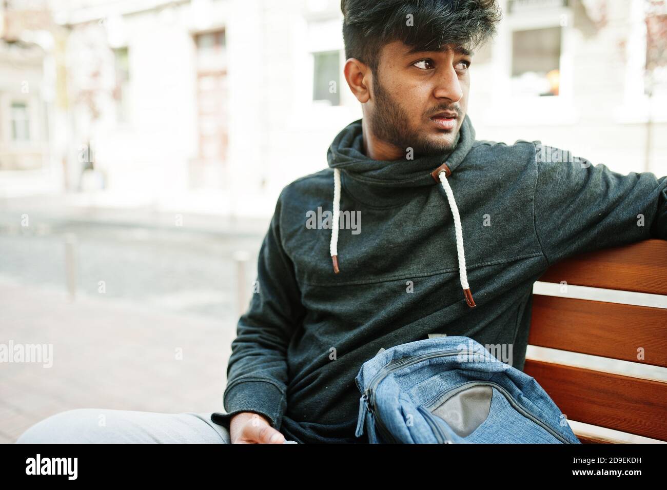 Portrait of young stylish indian man model pose in street, sitting on bench and hold smartphone ...