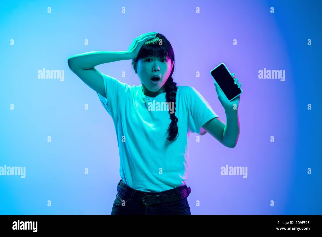 Shocked showing phone. Young asian woman's portrait on gradient blue ...
