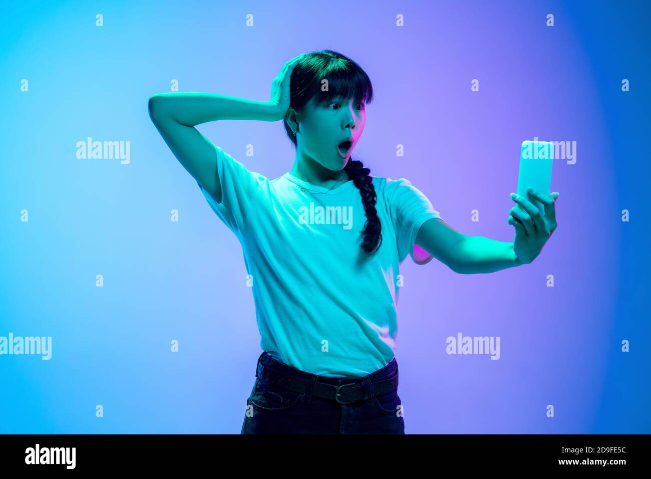 Shocked holding phone. Young asian woman's portrait on gradient blue ...