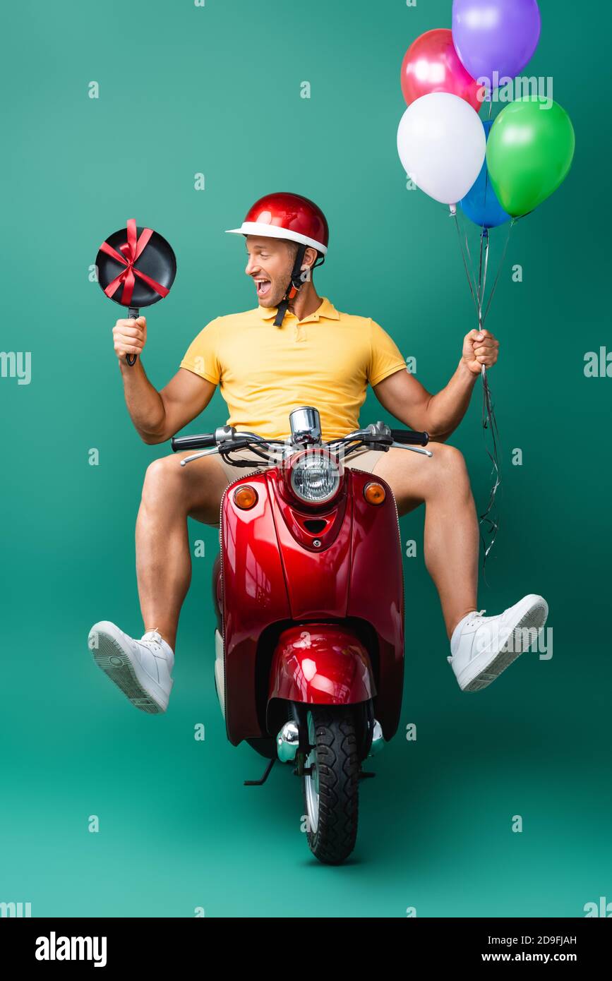 happy delivery man in helmet riding scooter while holding balloons and ...