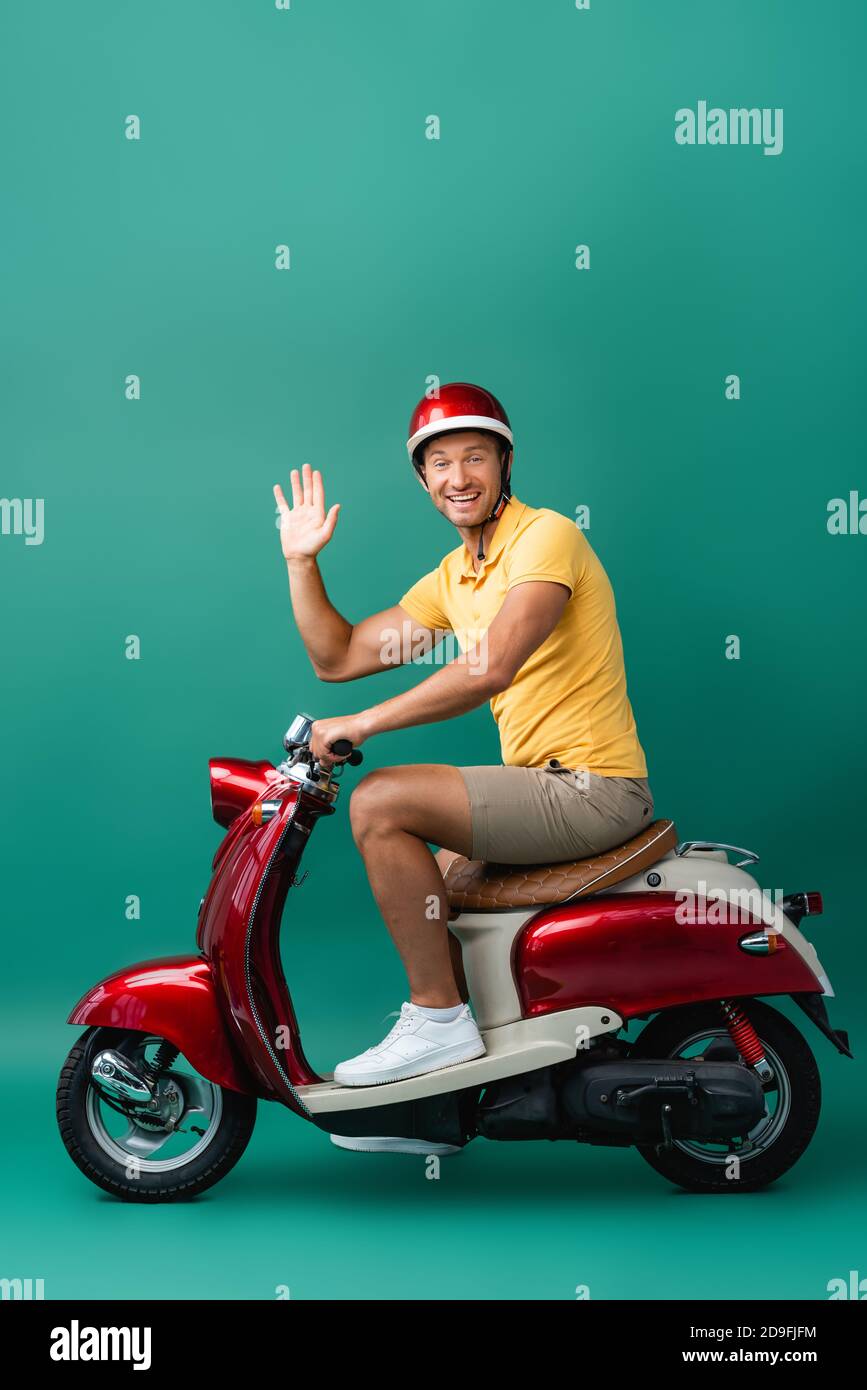 happy delivery man in helmet riding motorbike and waving hand on blue ...