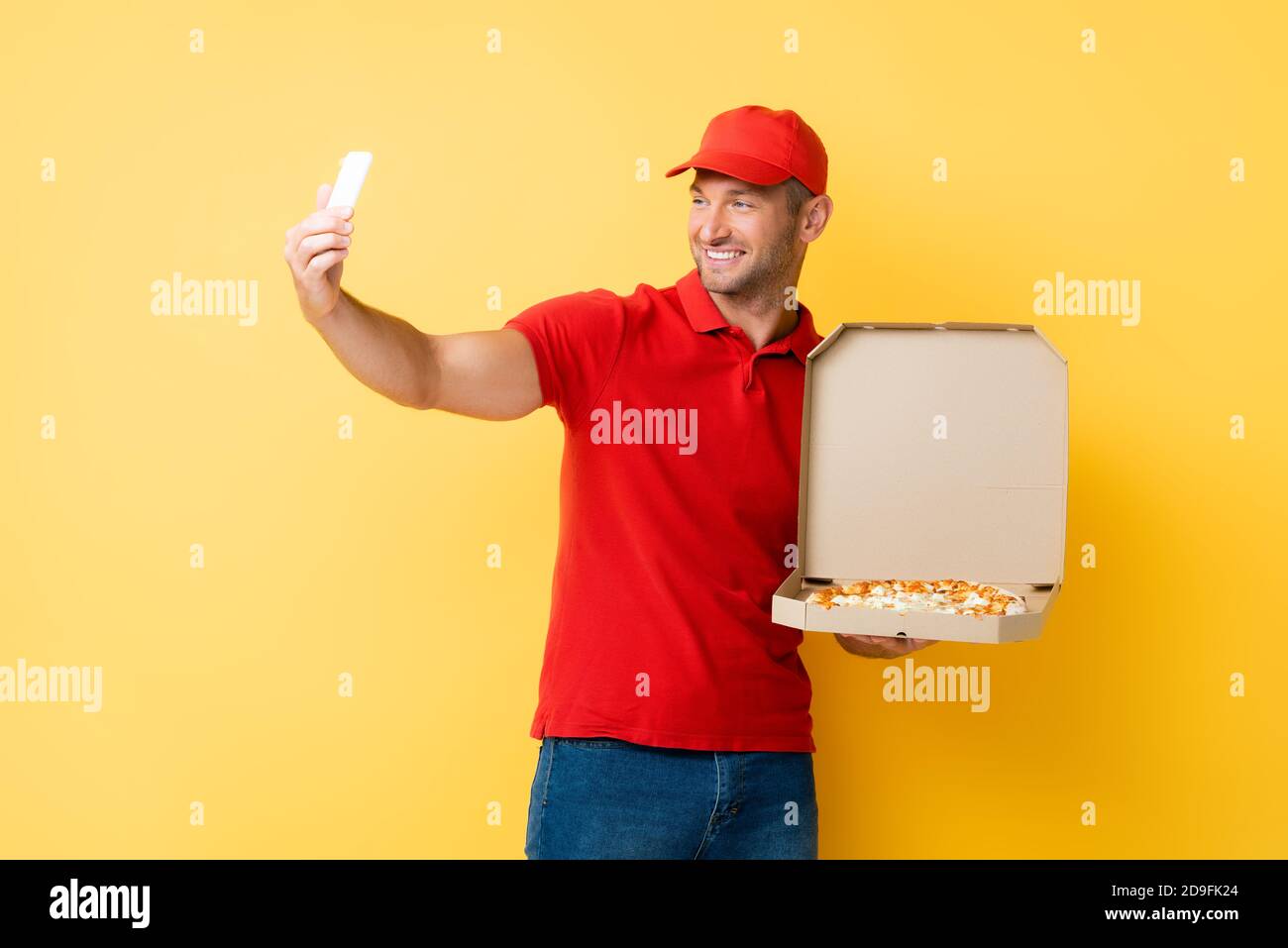 smiling delivery man in red cap holding box with tasty pizza and taking ...