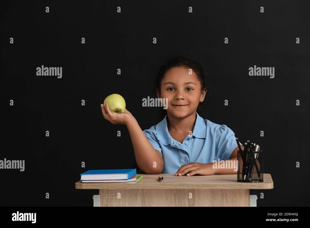 Little African-American pupil sitting at school desk against dark ...