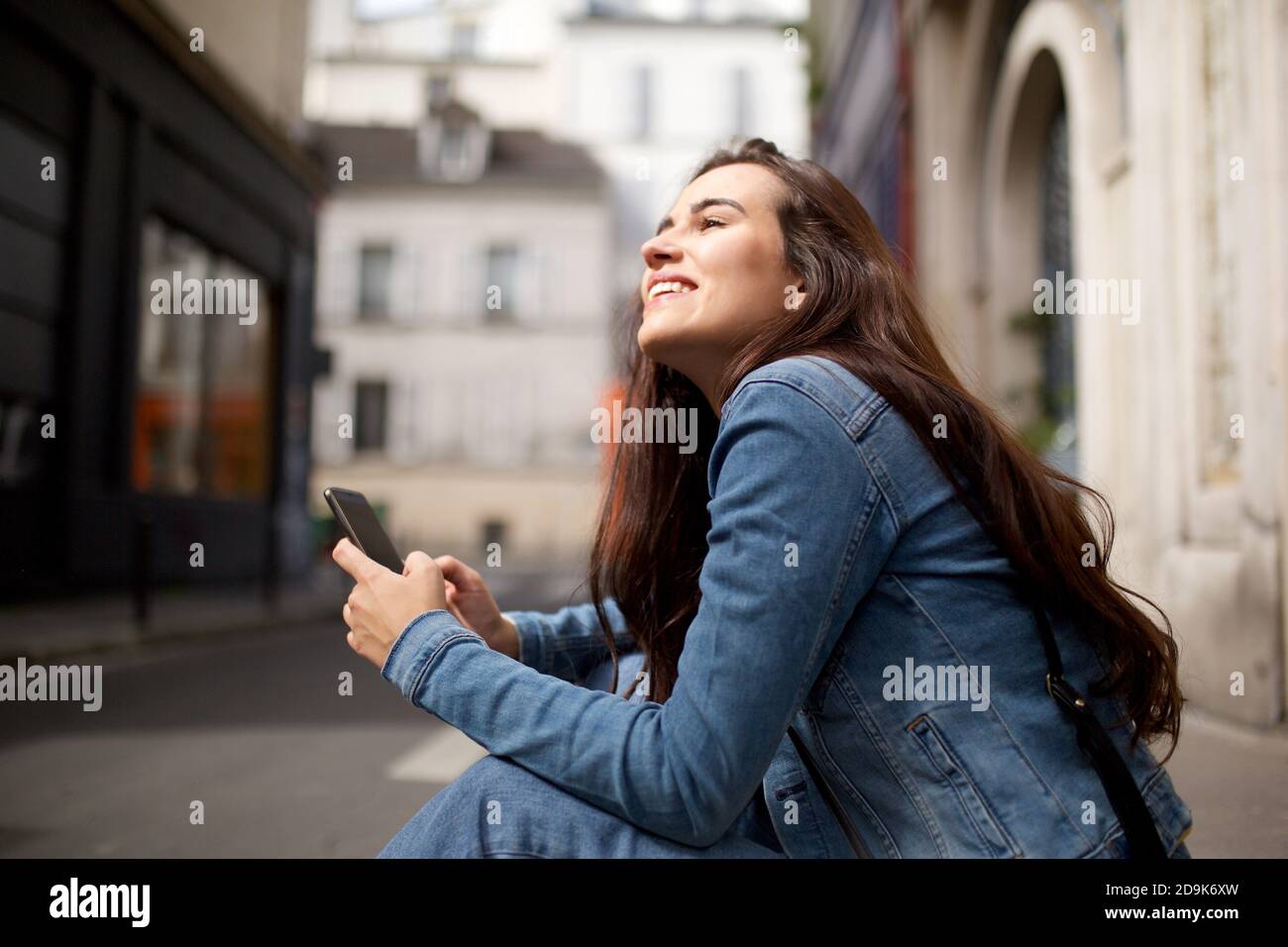 Side portrait smiling young woman sitting outside holding mobile phone ...