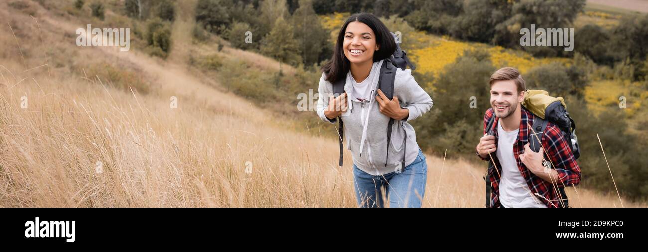 Cheerful multicultural hikers walking with backpacks on field, banner ...