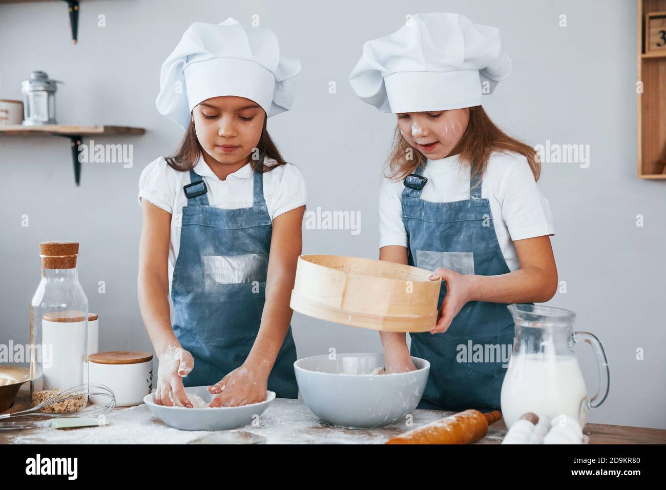 Two little girls in blue chef uniform working with flour by using sieve ...