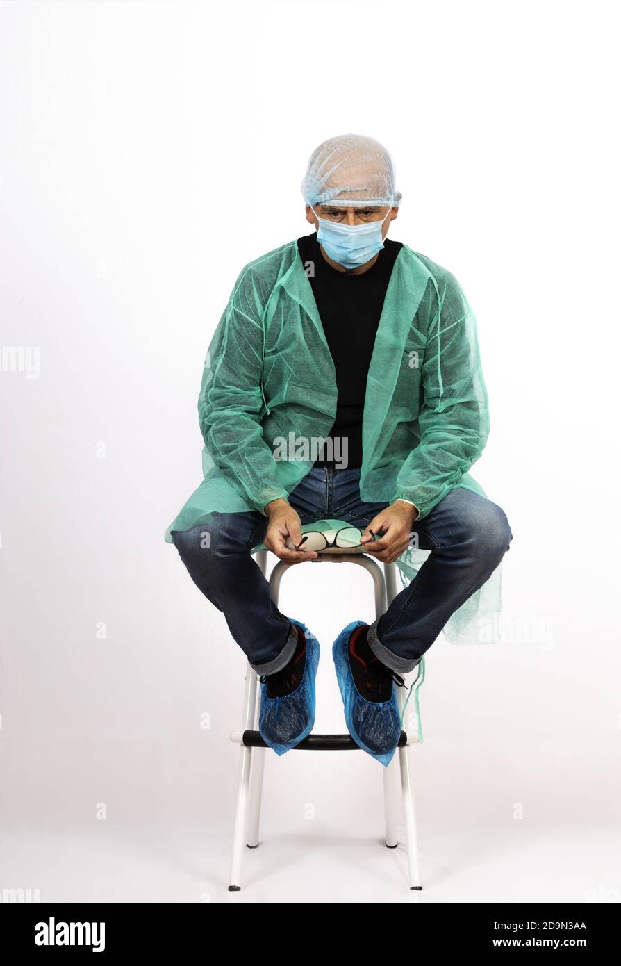 Man wearing a face mask, surgical gown, cap and shoe covers and sitting ...