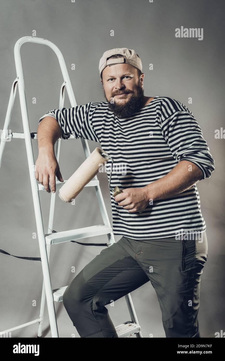 Joyful bearded craftsman with brush roller and ladder studio portrait ...