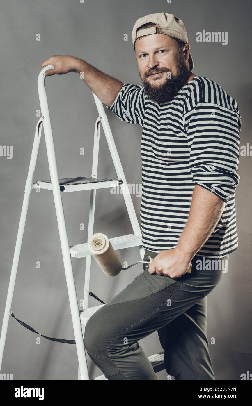 Joyful bearded craftsman with brush roller and ladder studio portrait ...