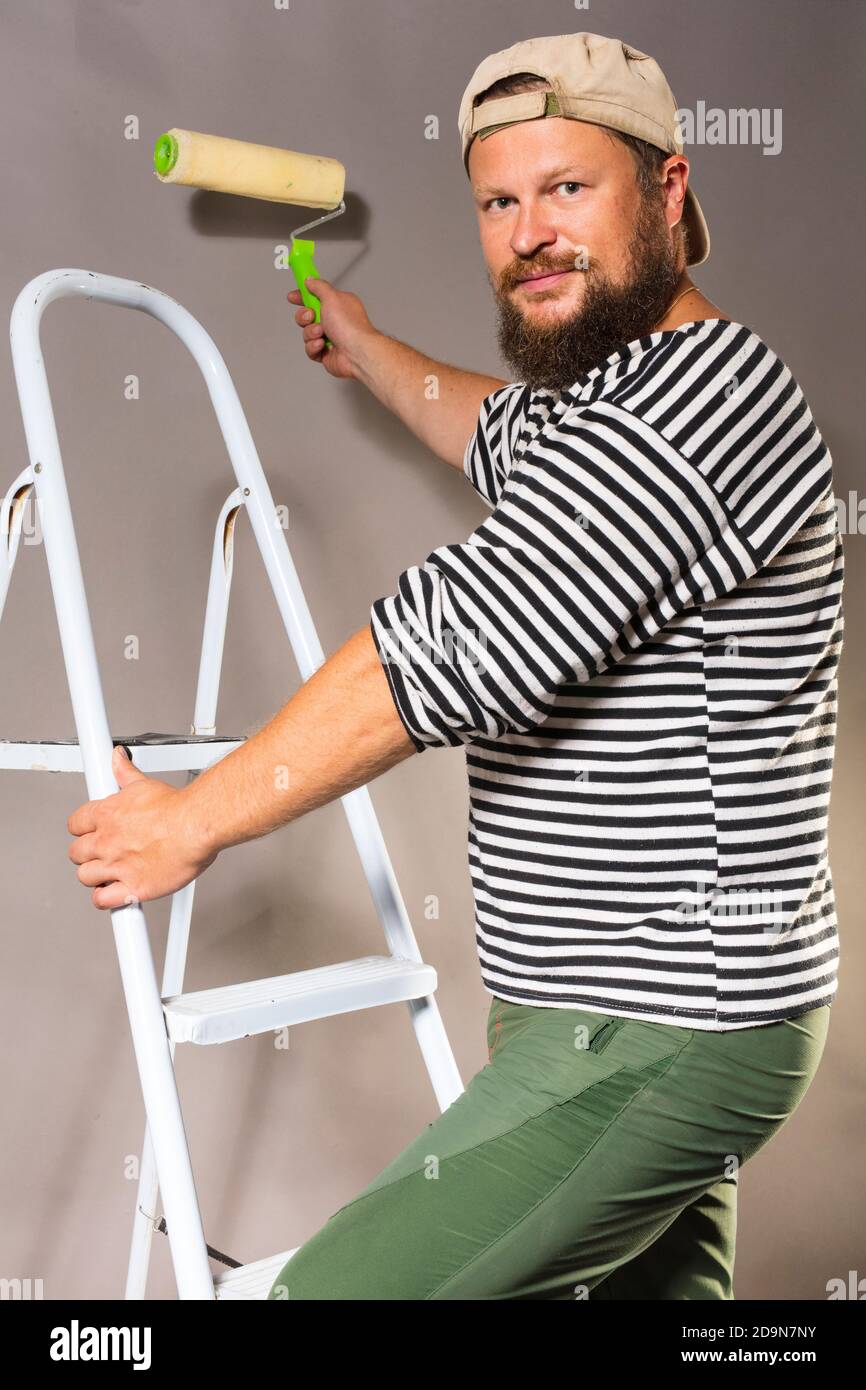 Joyful bearded craftsman with brush roller and ladder studio portrait ...
