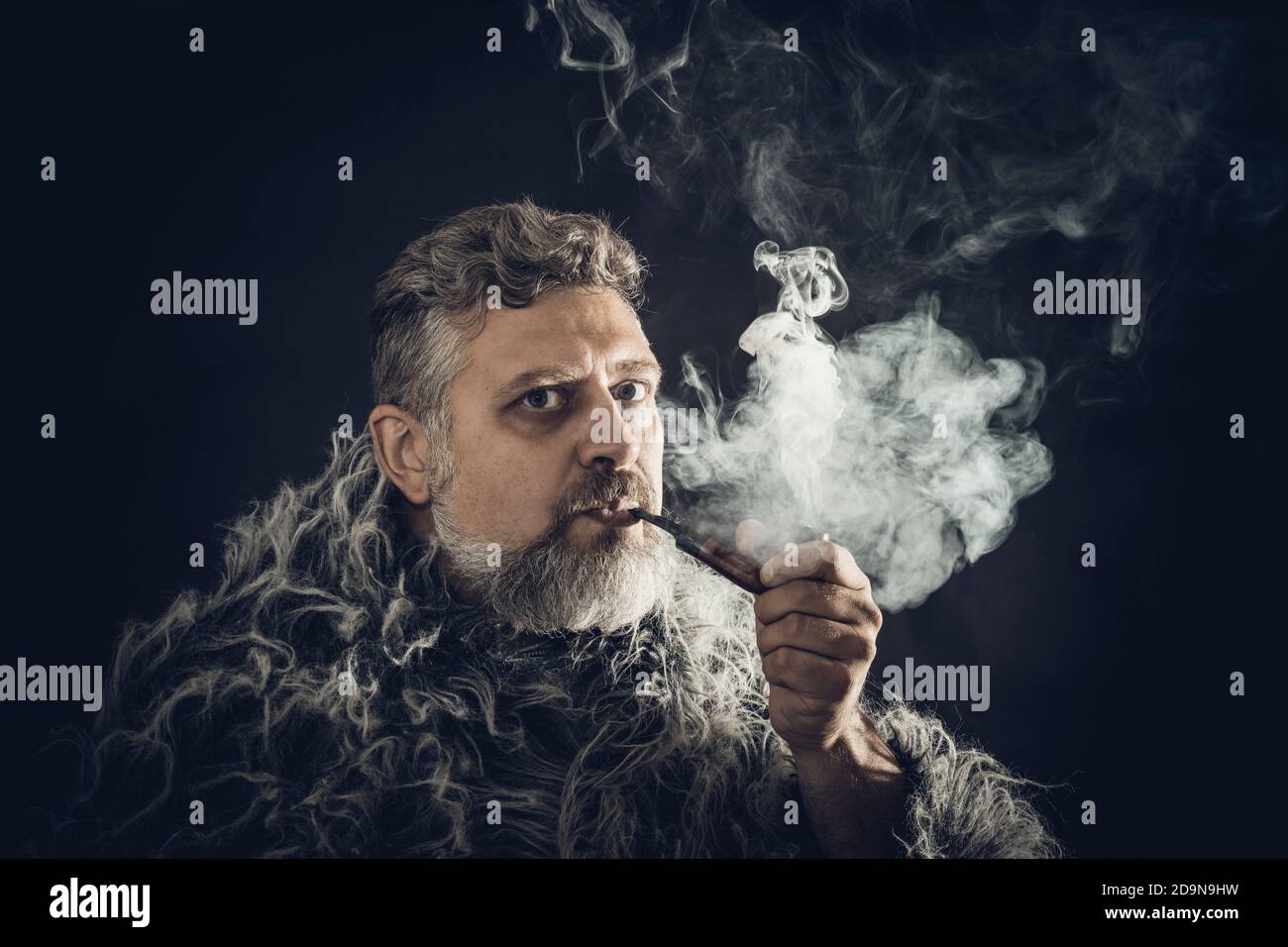 Solid bearded man in a fur mantle smoking pipe studio portrait on black ...