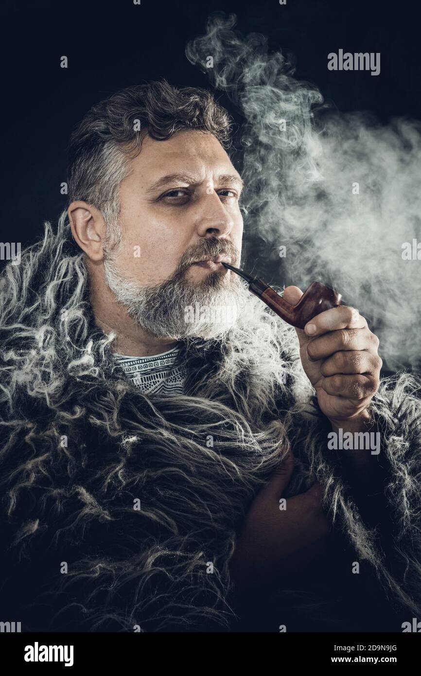 Solid bearded man in a fur mantle smoking pipe studio portrait on black ...