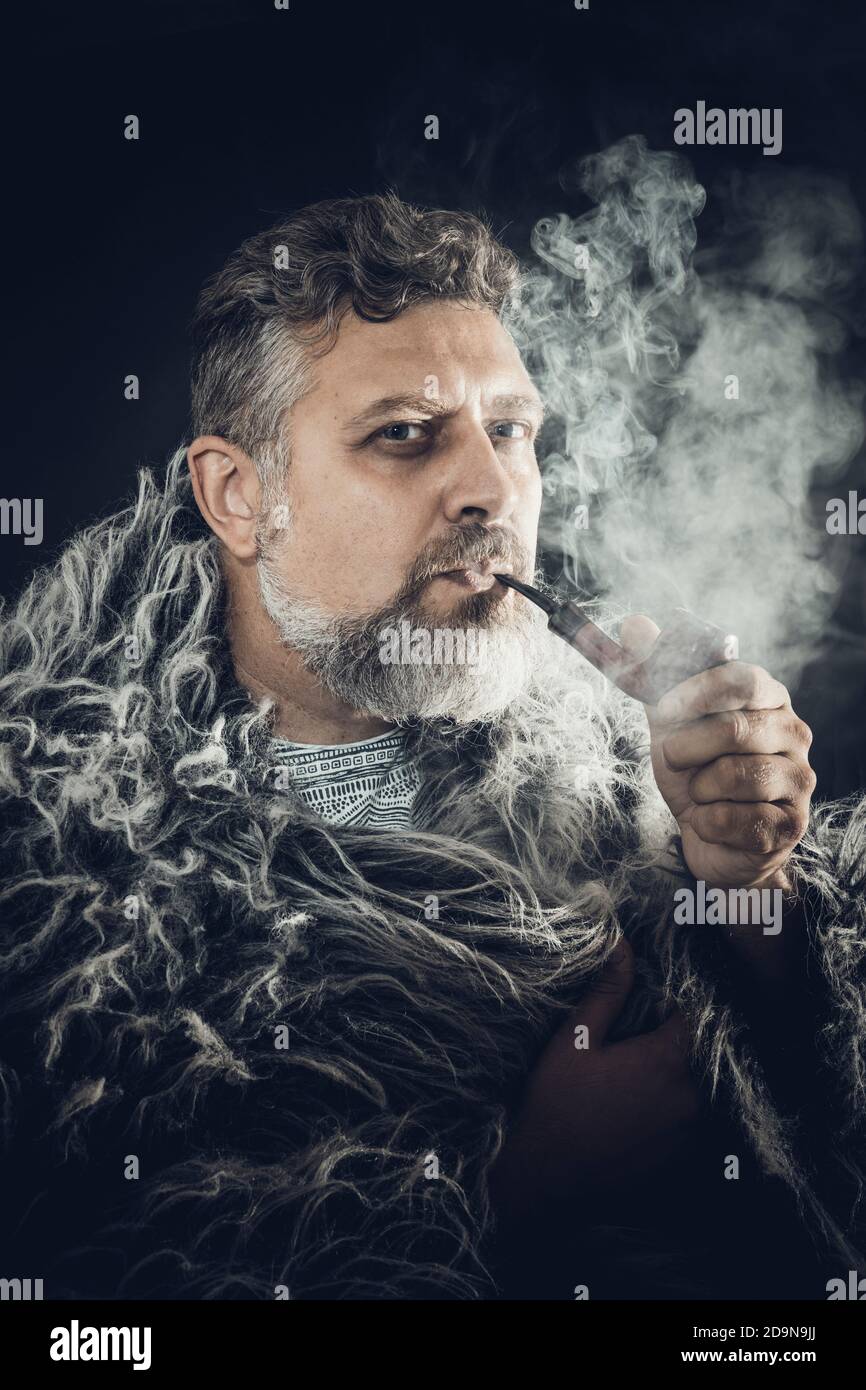 Solid bearded man in a fur mantle smoking pipe studio portrait on black ...