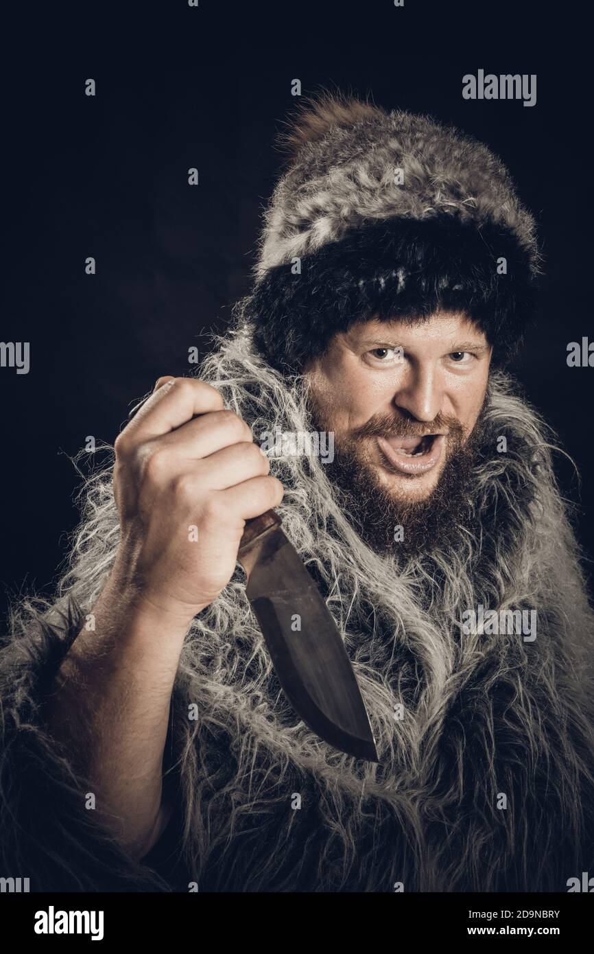 Bearded hunter dressed in a fur mantle and fur hat with knife studio ...