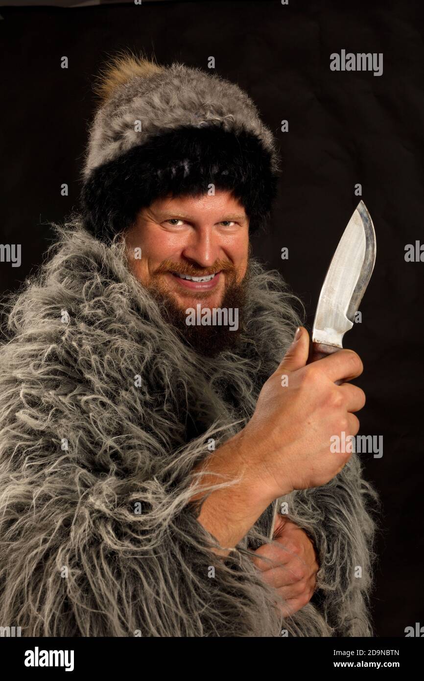 Bearded hunter dressed in a fur mantle and fur hat with knife studio ...