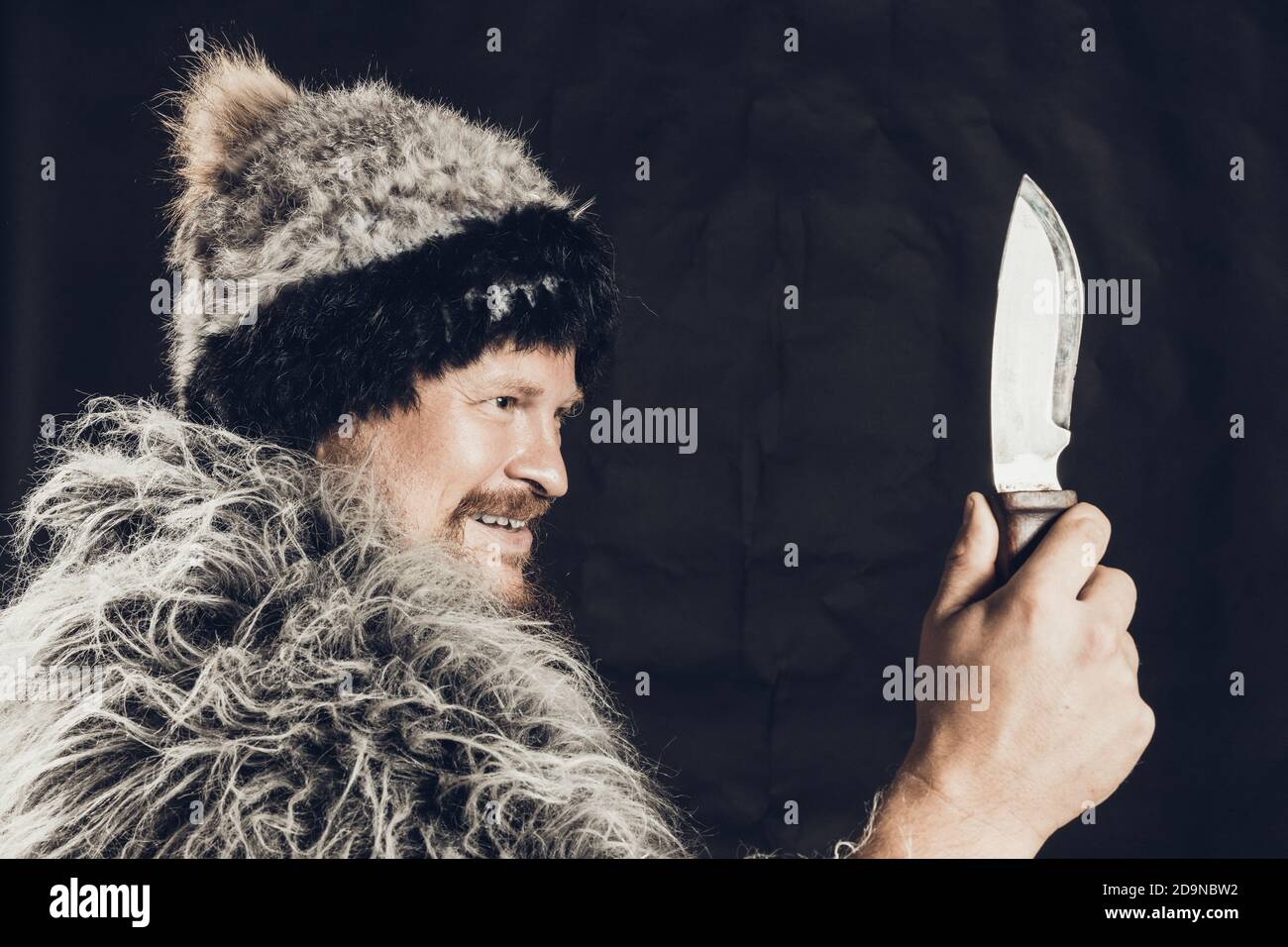 Bearded hunter dressed in a fur mantle and fur hat with knife studio ...