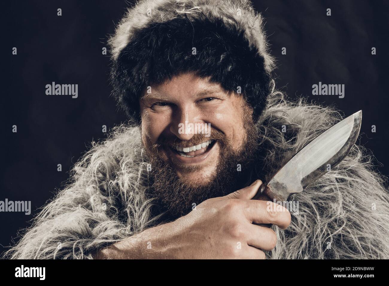 Bearded hunter dressed in a fur mantle and fur hat with knife studio ...