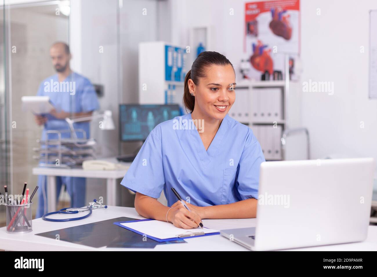 Smiling nurse using laptop computer and writing notes on clipboard in ...