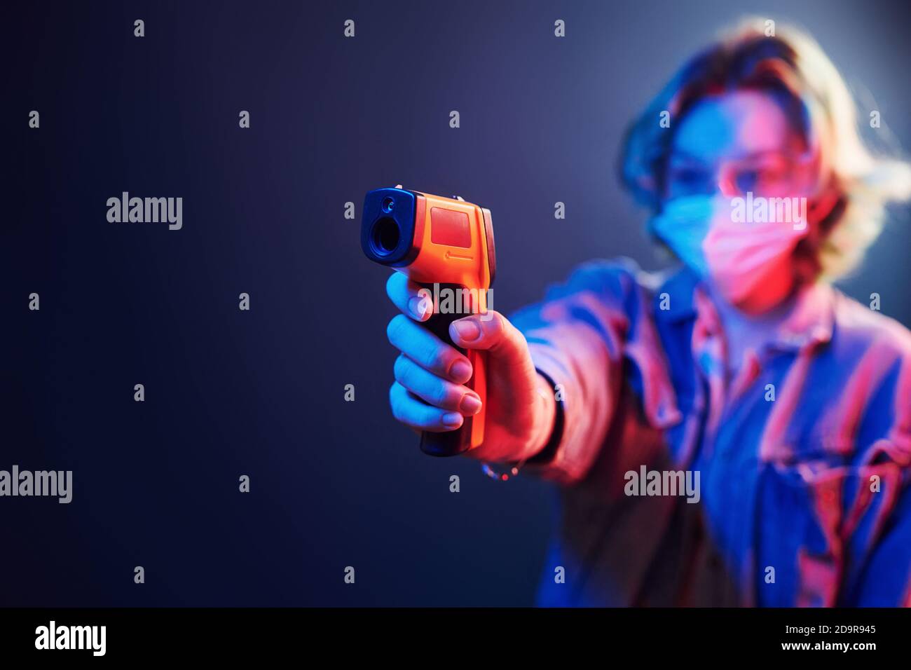 Girl in protective glasses and mask holding infrared thermometer. Neon ...