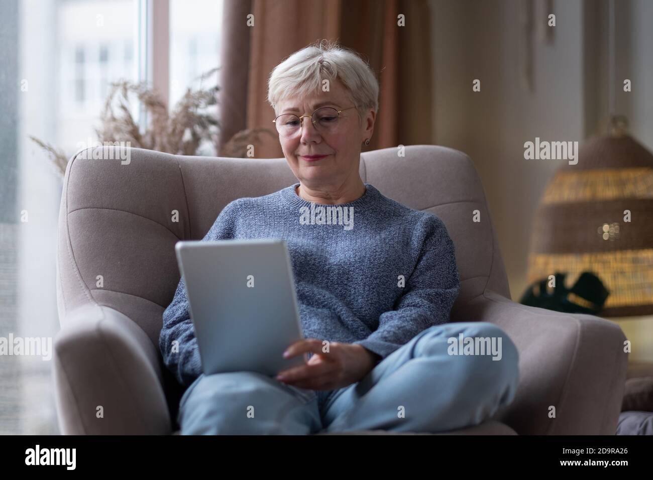 Senior woman with tablet pc resting on sofa near window reading ...
