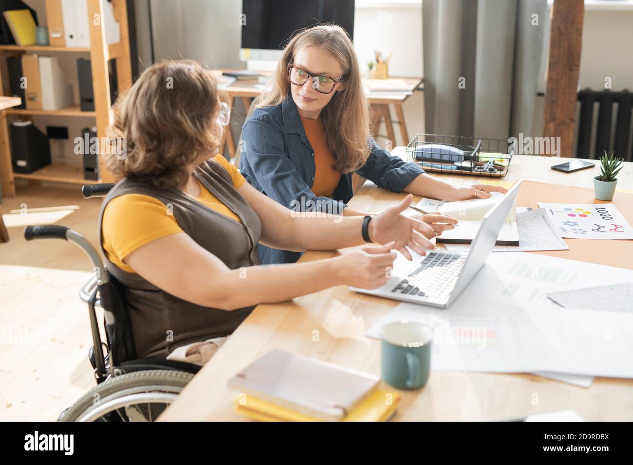 Two young female office managers sitting by table and discussing ...
