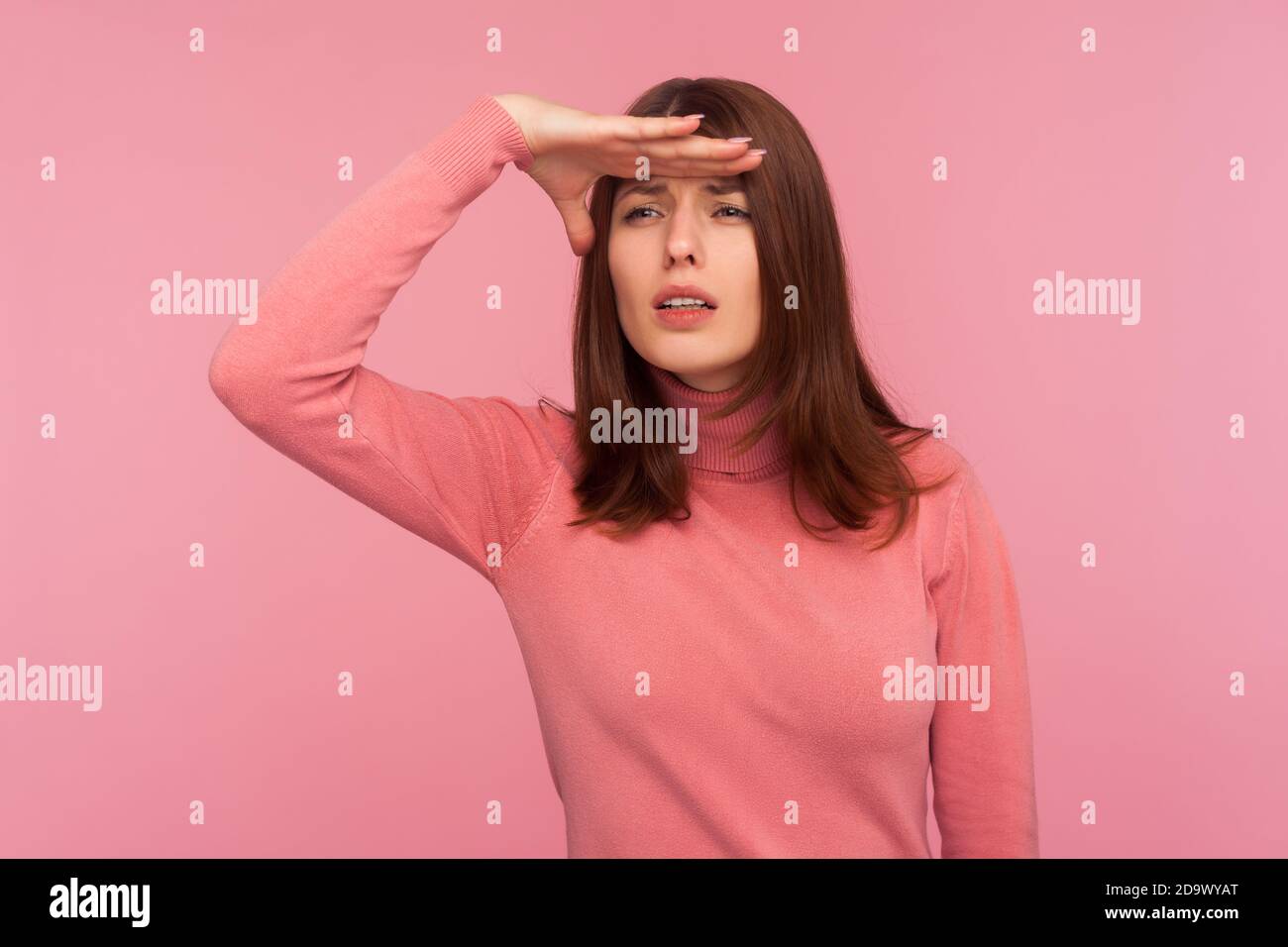 Curious brunette woman in pink sweater looking far away with hand over ...