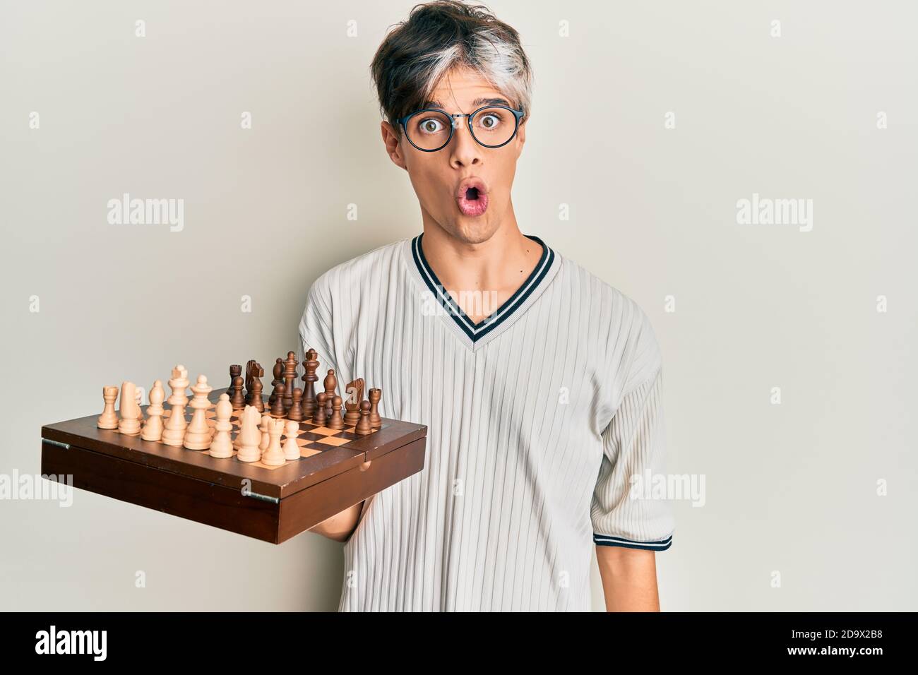 Young hispanic man holding chess scared and amazed with open mouth for ...