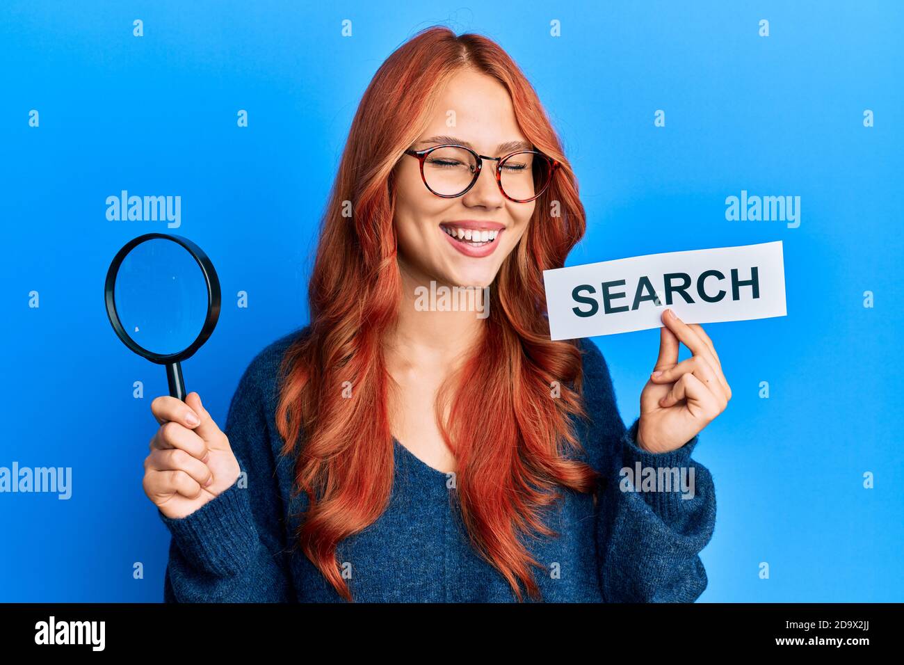 Young redhead woman holding magnifying glass and search word smiling ...