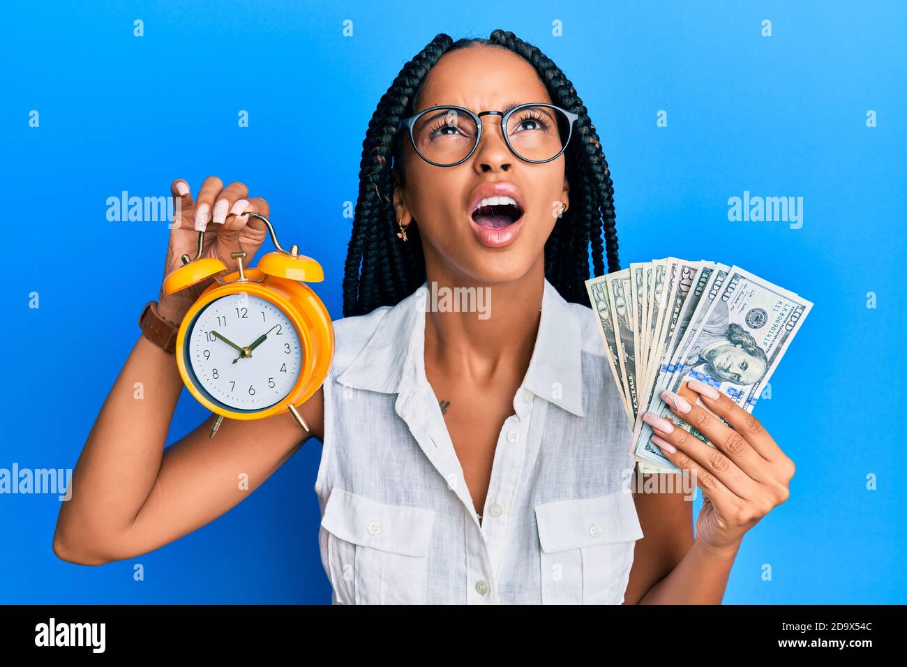 Beautiful hispanic woman holding alarm clock and dollars angry and mad ...