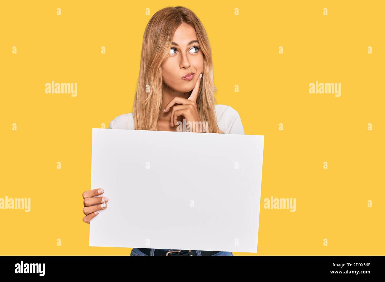 Beautiful blonde young woman holding blank empty banner serious face ...