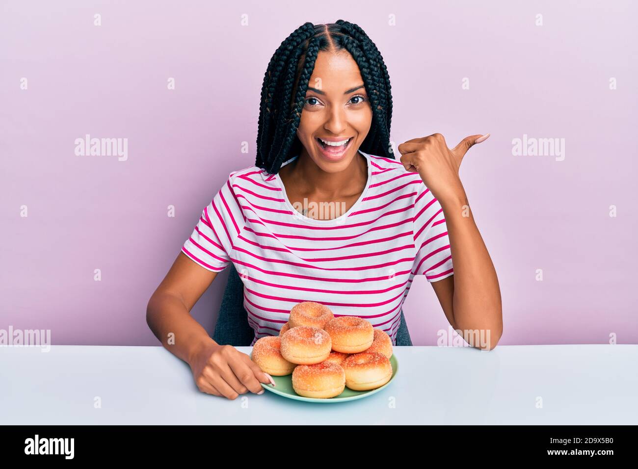 Beautiful hispanic woman eating doughnut for breakfast pointing thumb ...