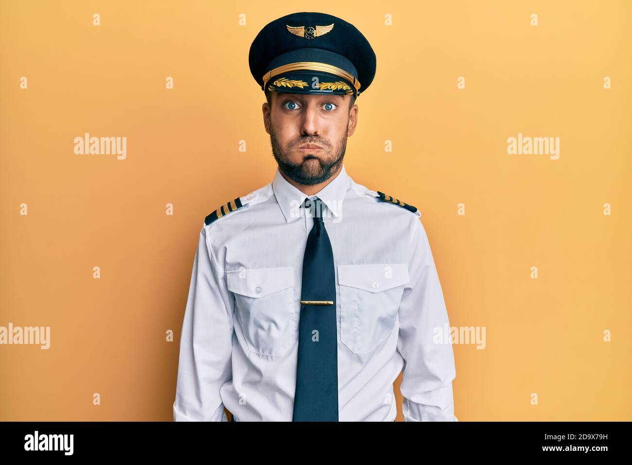 Handsome hispanic man wearing airplane pilot uniform puffing cheeks ...