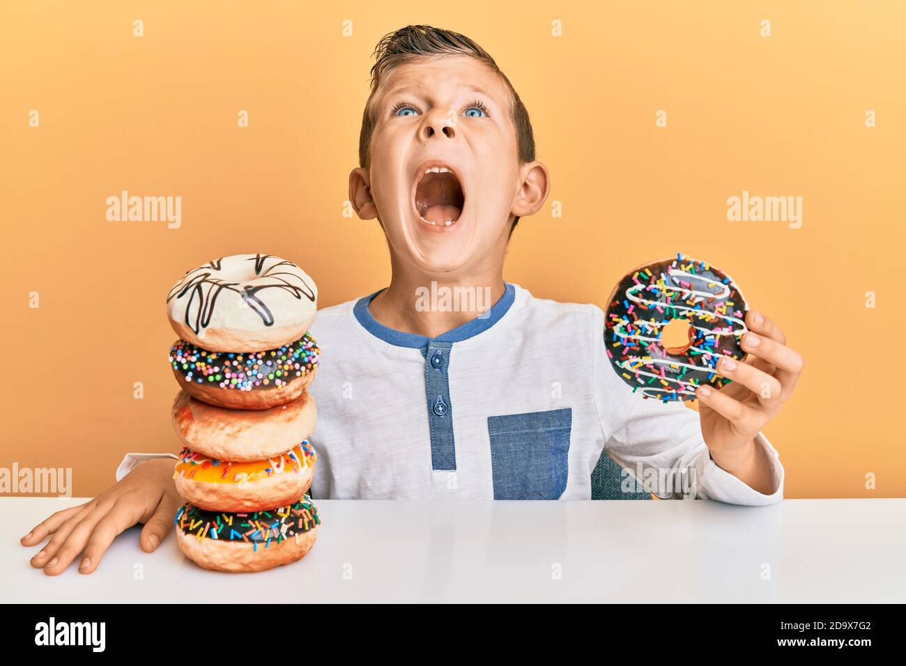 Adorable caucasian kid eating doughnuts for breakfast angry and mad ...