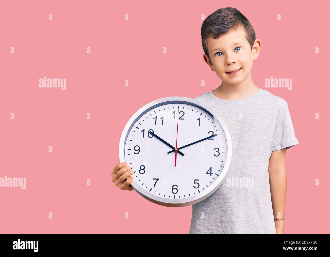 Cute blond kid holding big clock looking positive and happy standing ...