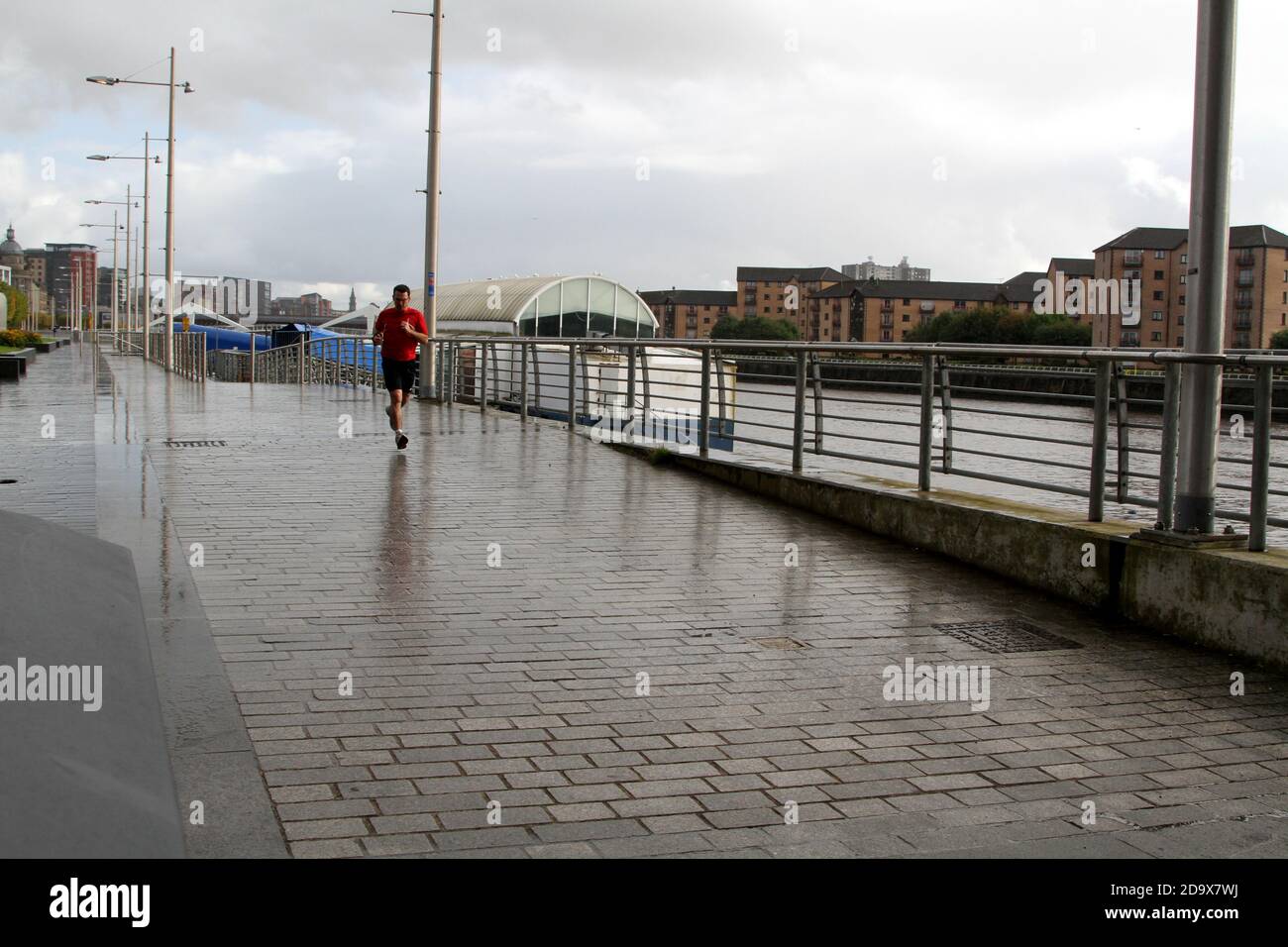 Glasgow, Scotland, UK. A jogger in red shirt runs in the rain alongside the River Clyde in the ...